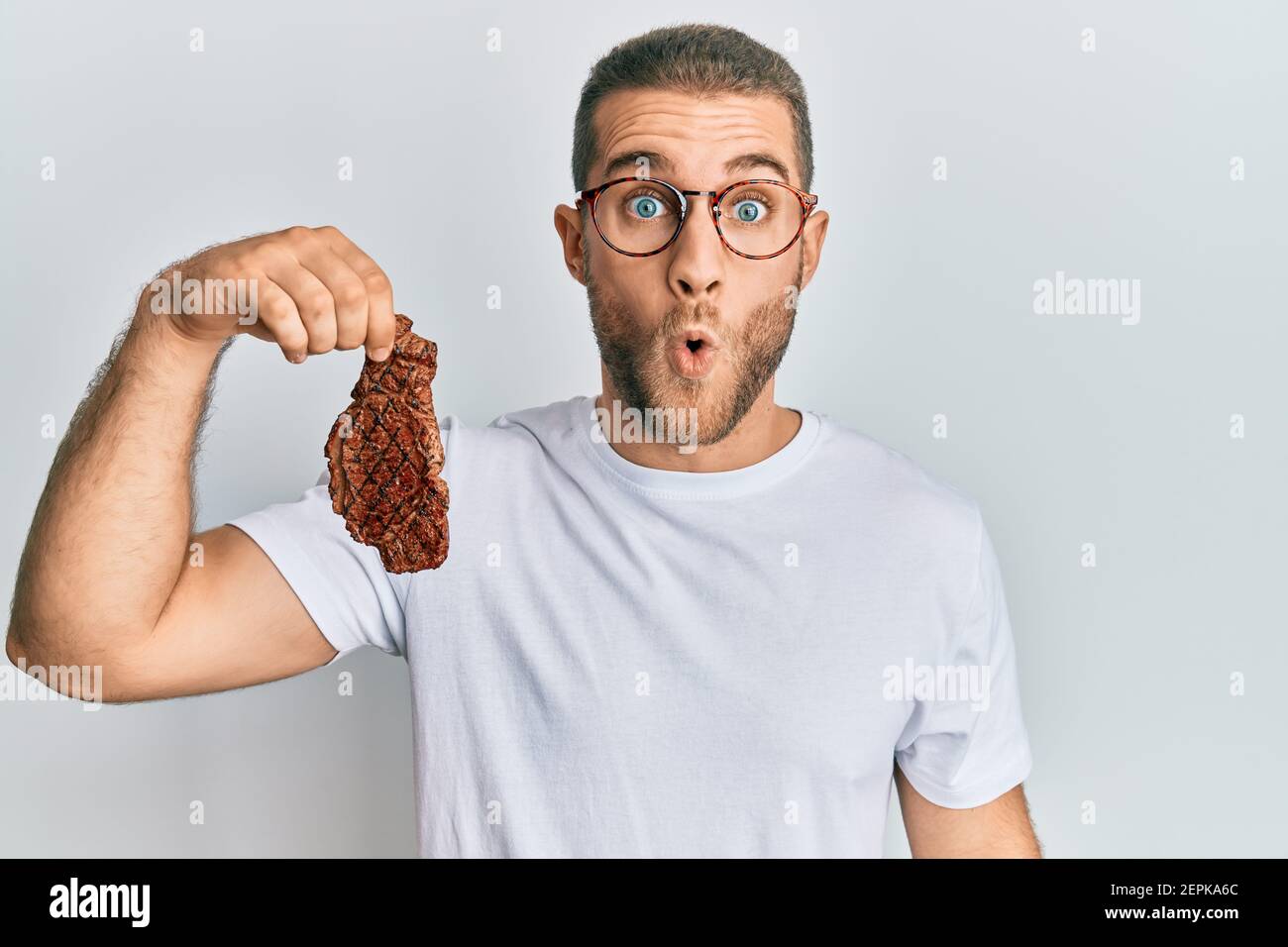 Young caucasian man holding beef steak scared and amazed with open ...