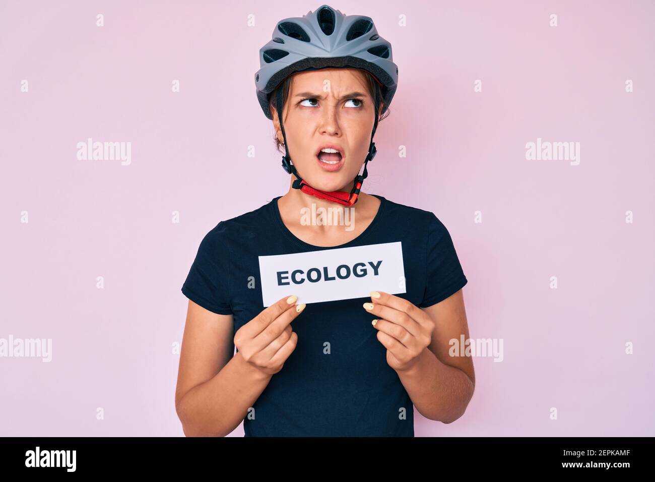 Beautiful caucasian woman wearing bike helmet holding ecology word angry and mad screaming ...
