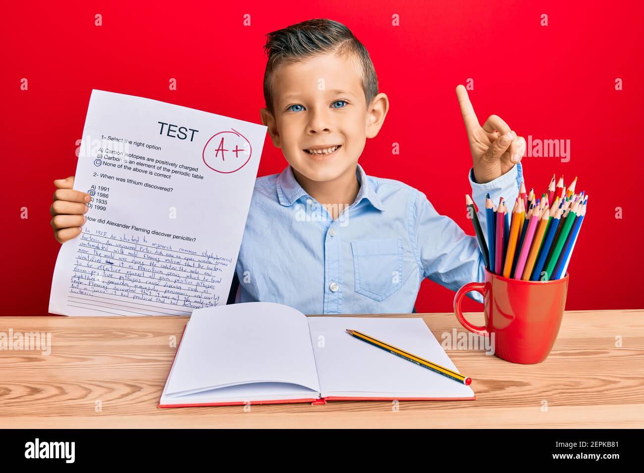 Adorable caucasian kid holding passed test sitting on the table smiling ...