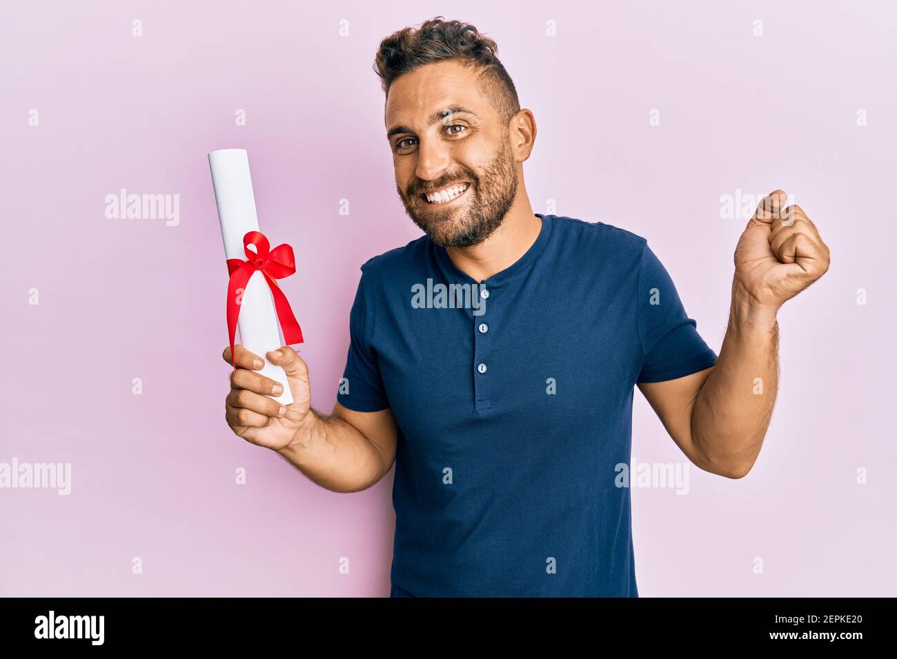 Handsome man with beard holding graduate degree diploma screaming proud ...