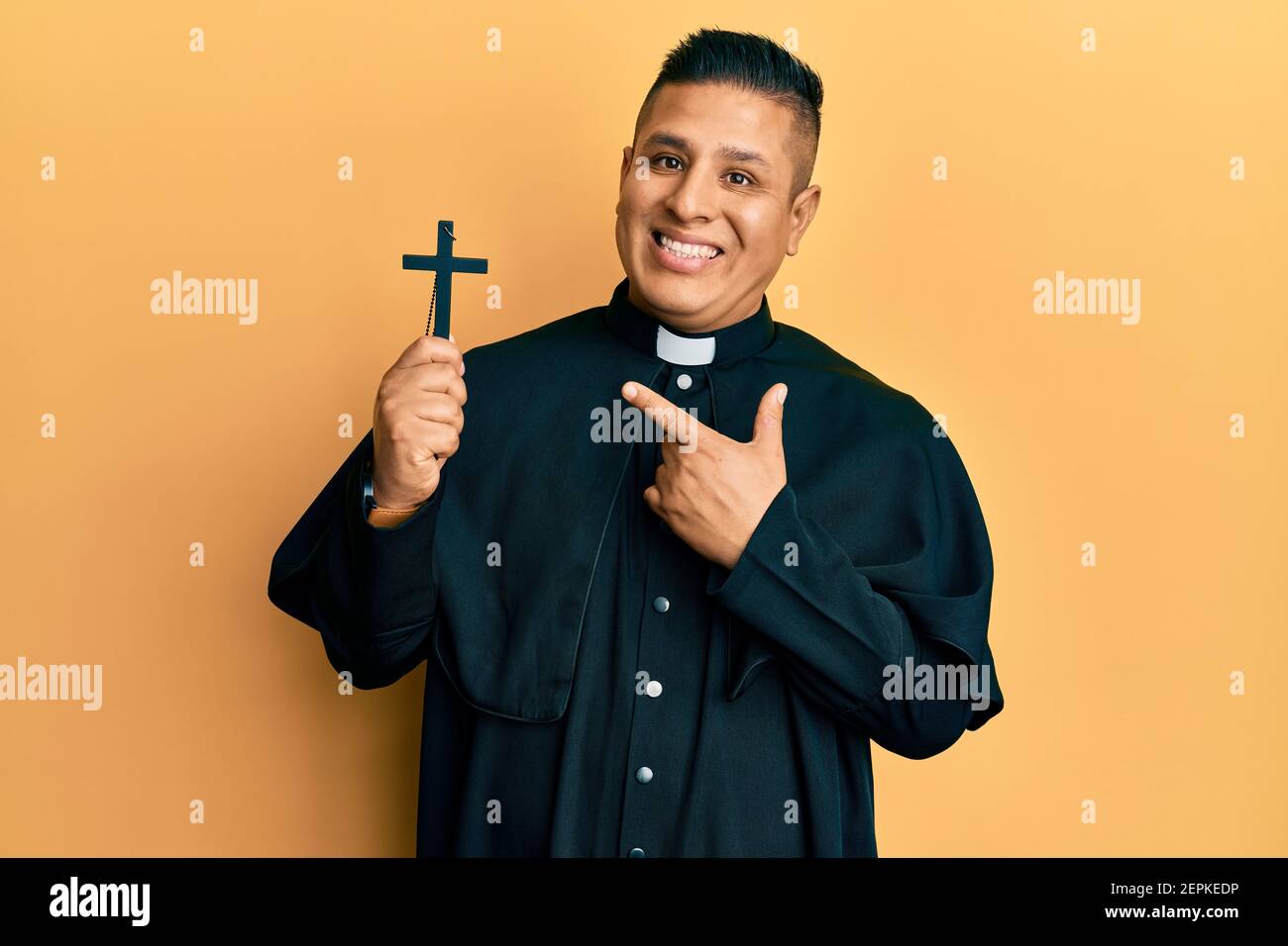 Young latin priest man holding crucifix smiling happy pointing with ...