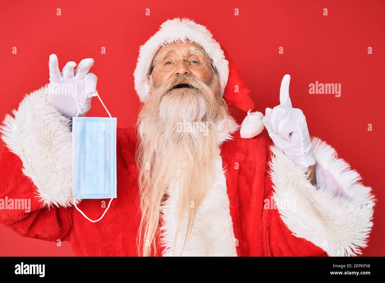 Old senior man wearing santa claus costume holding safety mask smiling ...