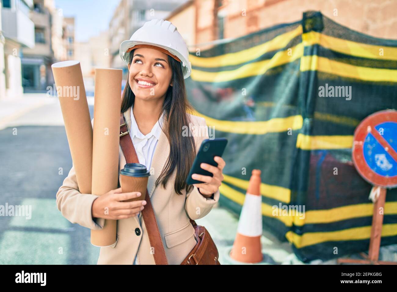 Young latin architect girl holding blueprints and using smartphone at ...