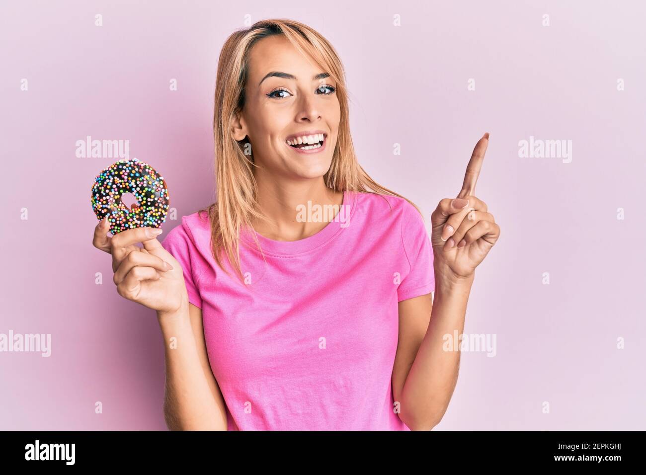 Beautiful blonde woman holding donut surprised with an idea or question ...