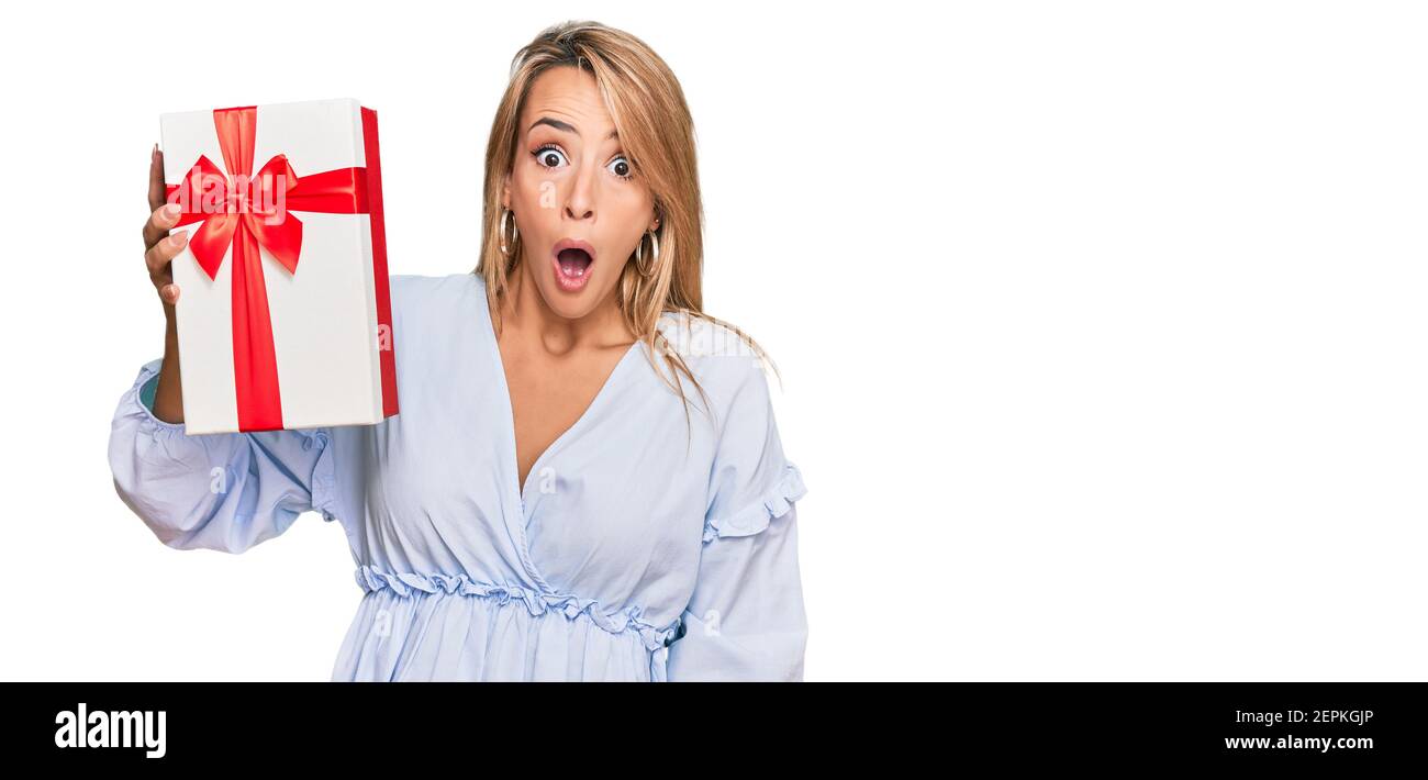 Beautiful blonde woman holding gift scared and amazed with open mouth ...