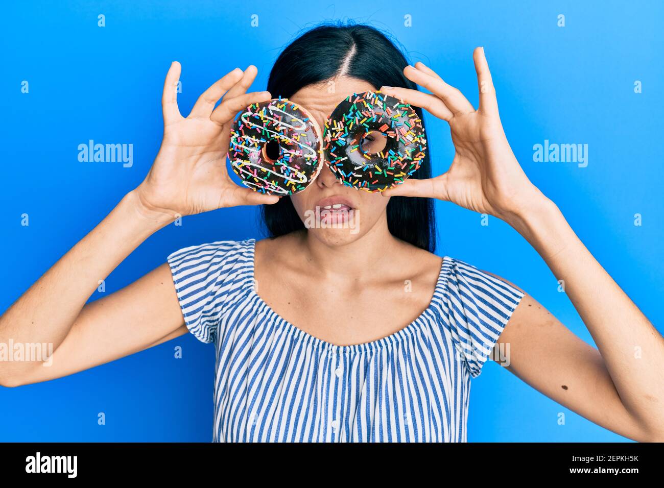 Beautiful young woman holding tasty colorful doughnuts on eyes clueless ...