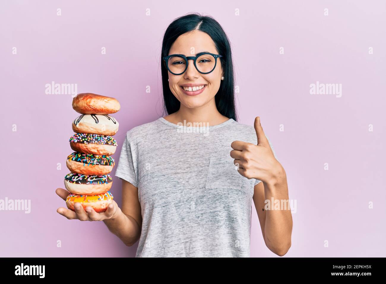 Beautiful young woman holding pile of tasty colorful doughnuts smiling ...