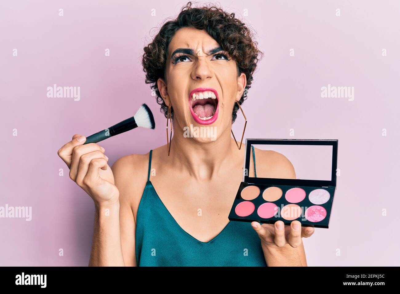 Young man wearing woman make up holding makeup brush and blush angry ...