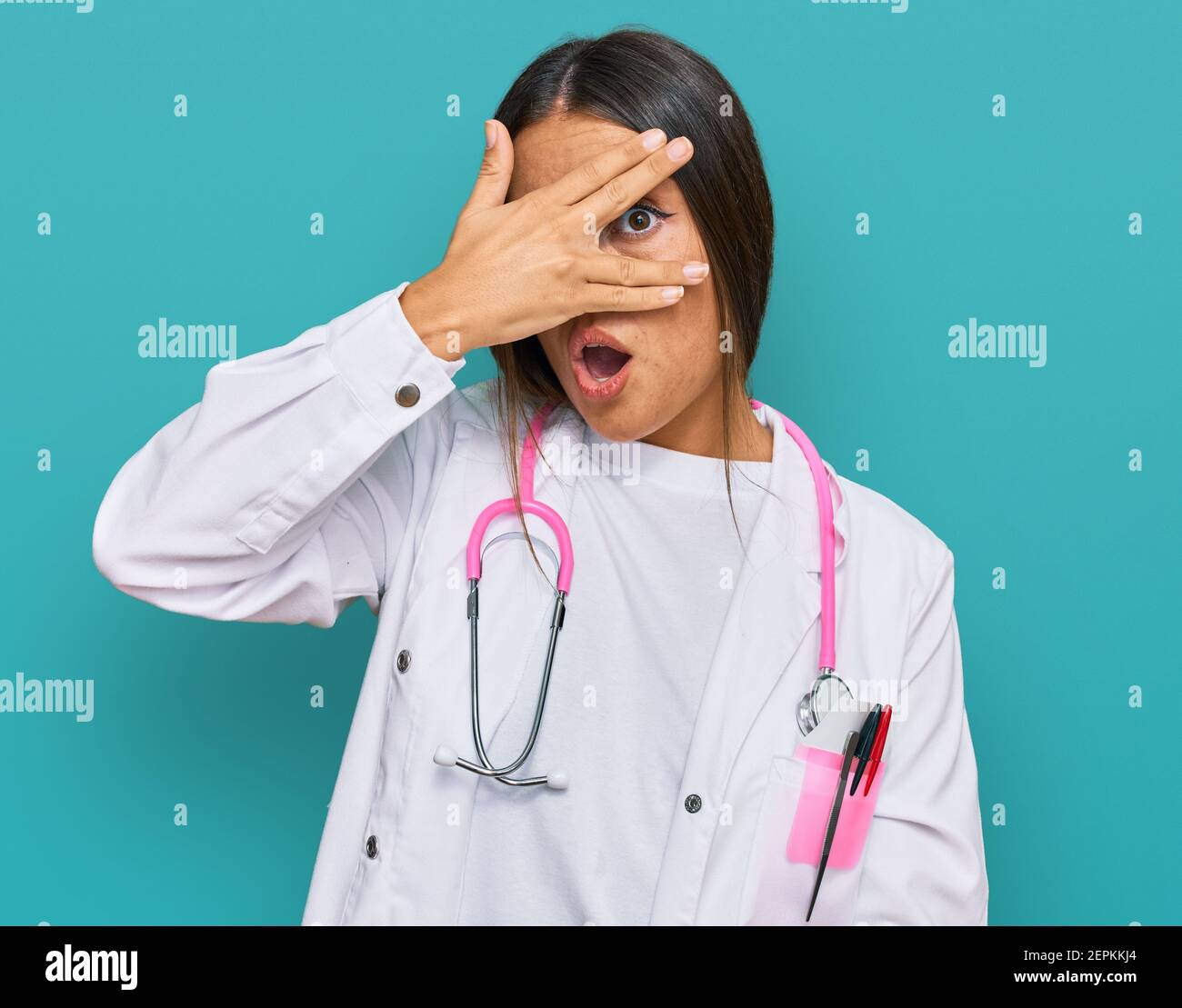 Beautiful hispanic woman wearing doctor uniform and stethoscope peeking ...