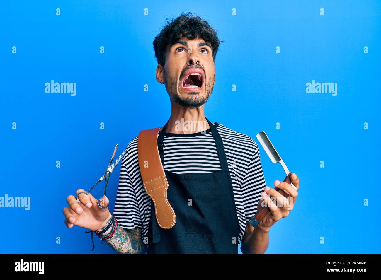 Young hispanic man wearing barber apron holding razor and scissors angry and mad screaming ...