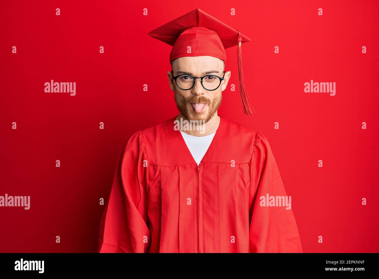 Young redhead man wearing red graduation cap and ceremony robe sticking ... Young redhead man wearing red graduation cap and ceremony robe sticking ...