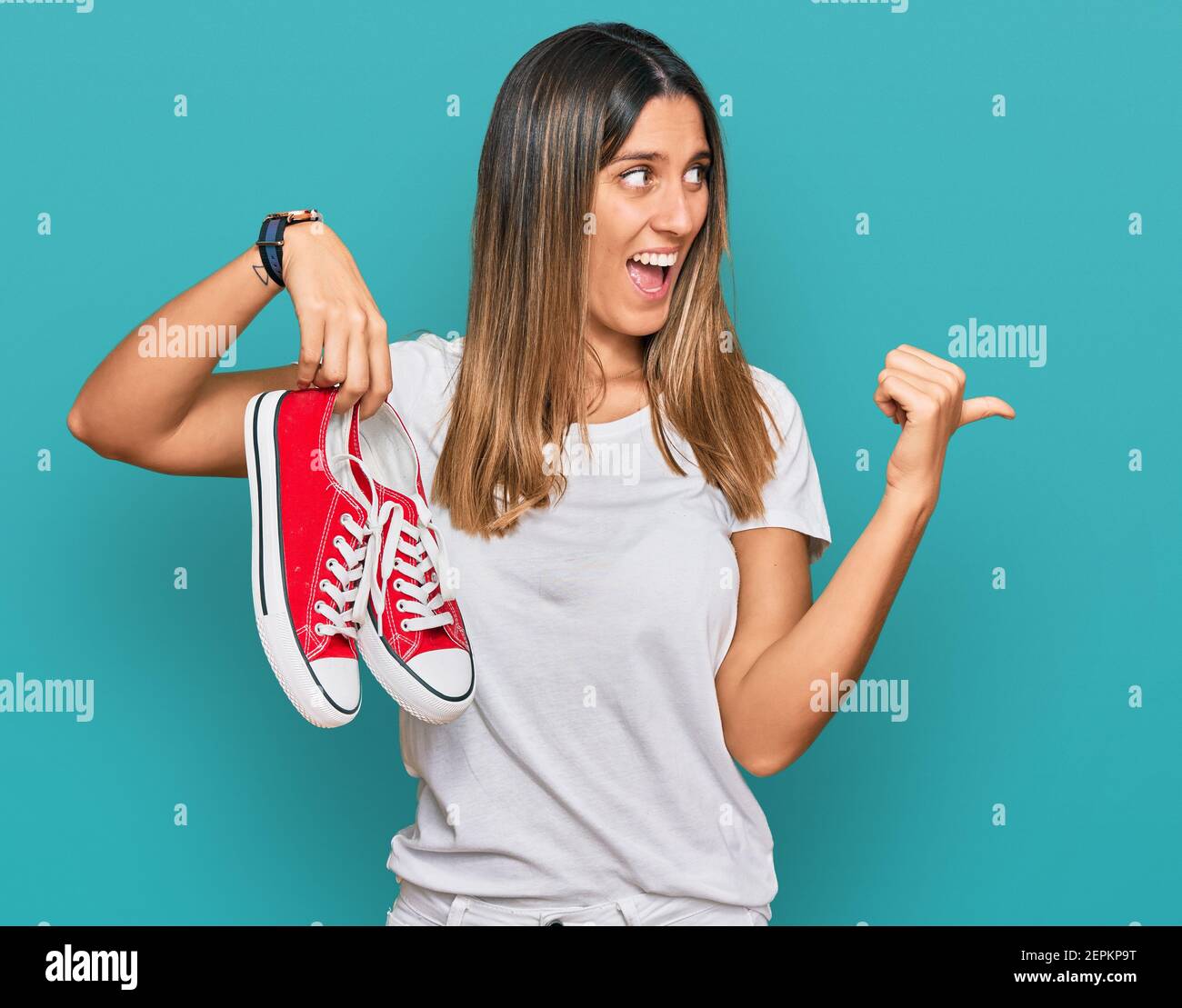 Young woman holding red casual shoes pointing thumb up to the side ...