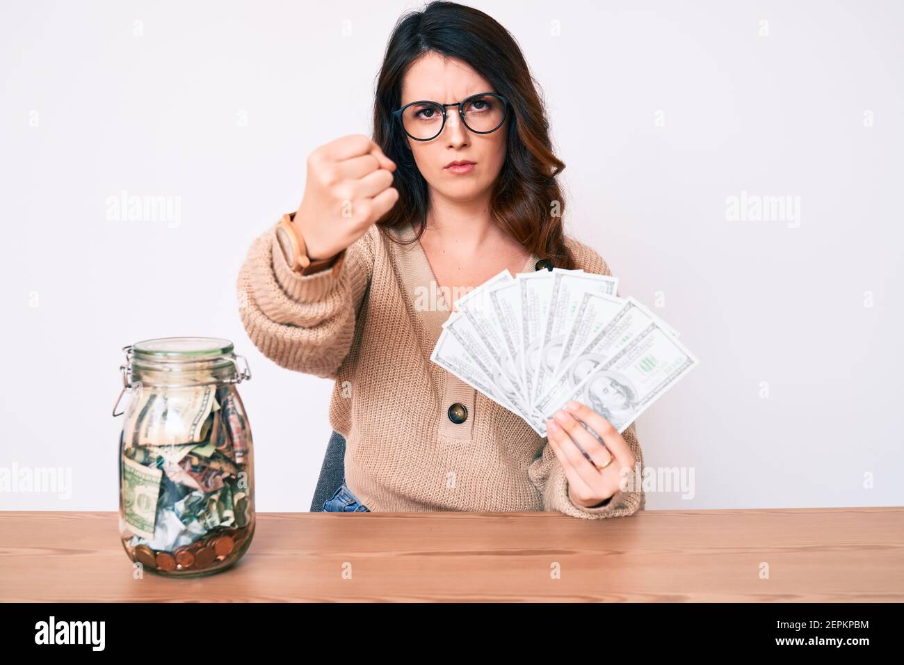 Young beautiful brunette woman holding jar with savings holding dollars ...