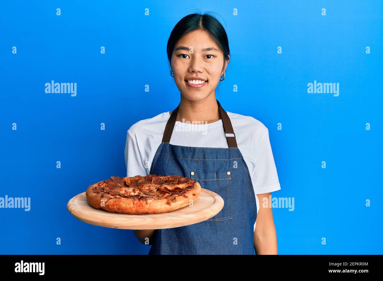 Young chinese woman wearing waiter apron holding italian pizza looking ...