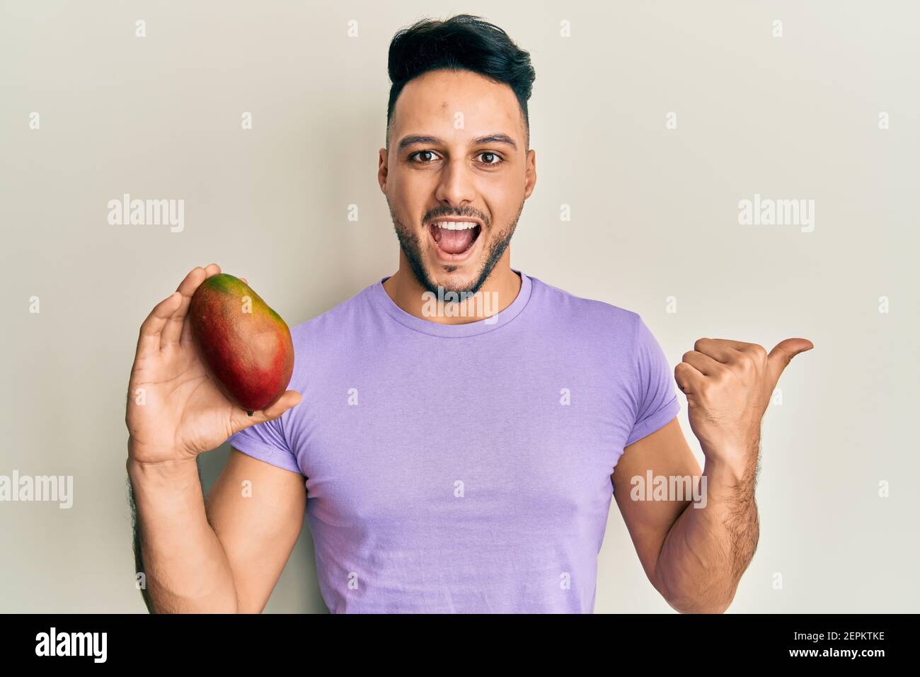 Young arab man holding mango pointing thumb up to the side smiling ...