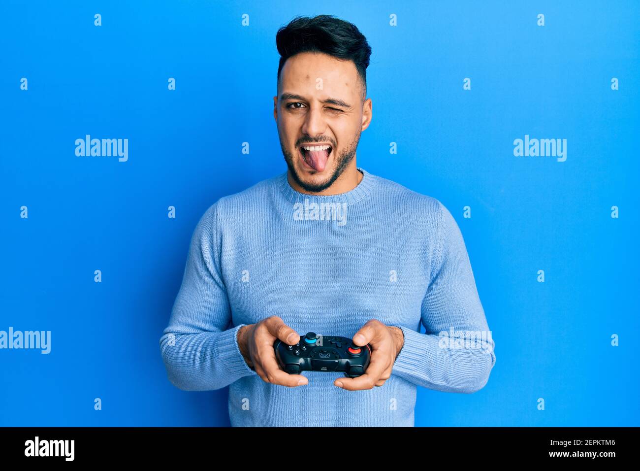 Young arab man playing video game holding controller sticking tongue ...