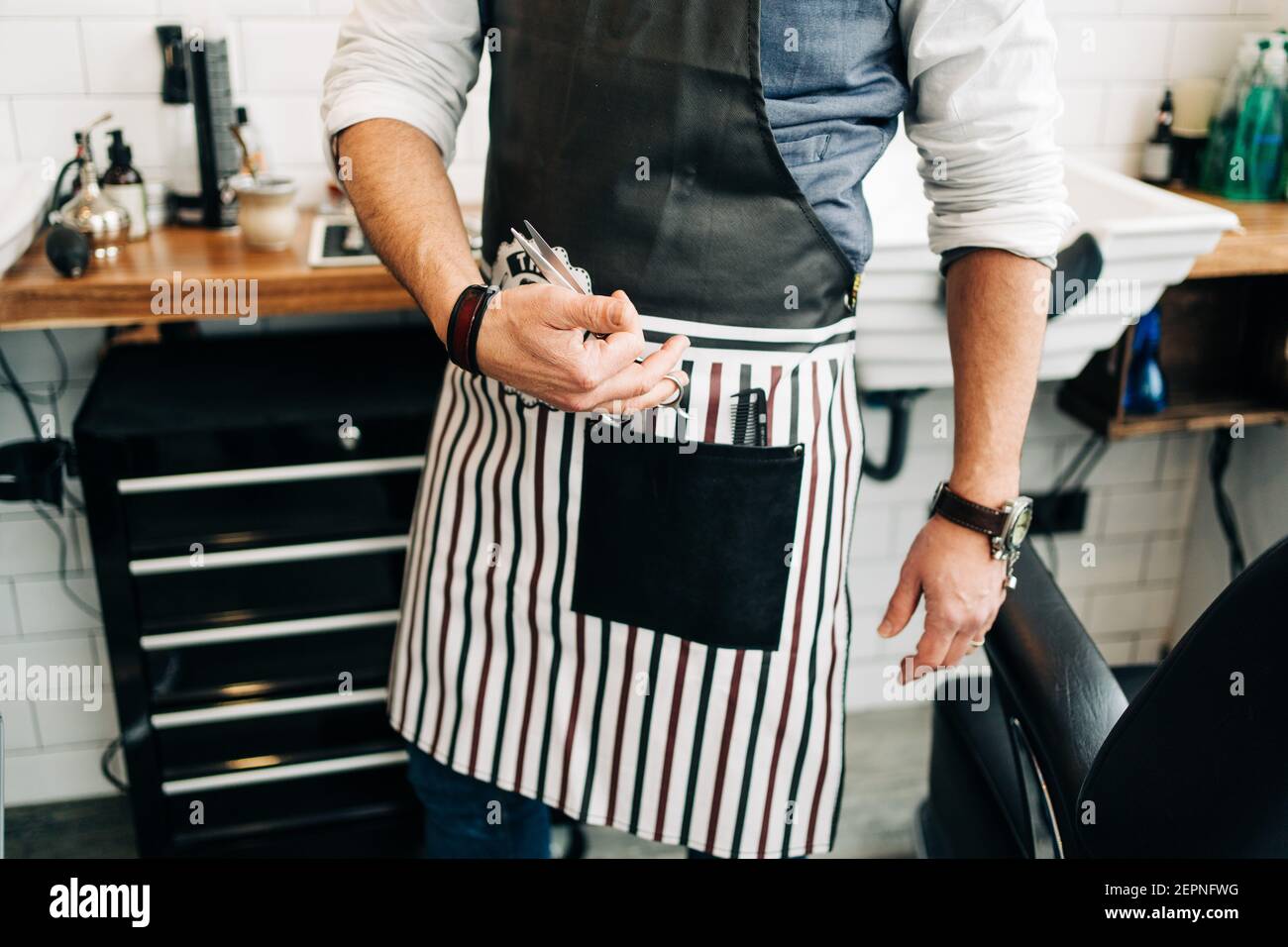 Crop unrecognizable male barber with scissors and comb in striped apron ...