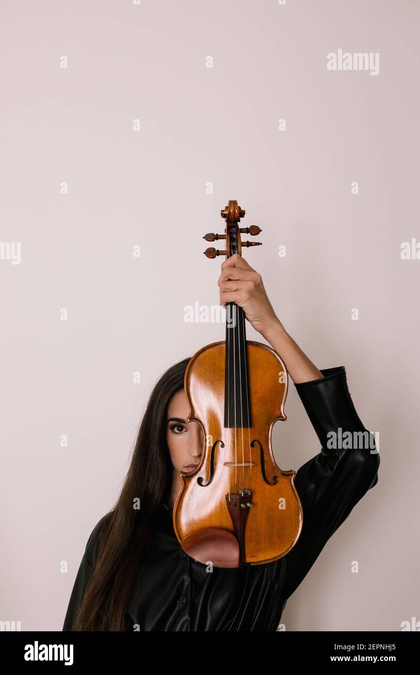 Female artist covering face with violin while standing against white ...