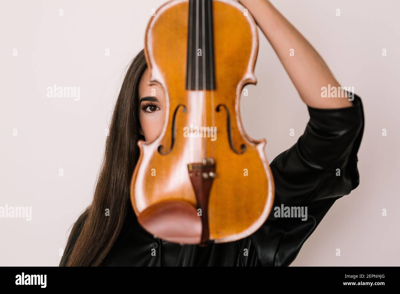 Female artist covering face with violin while standing against white ...