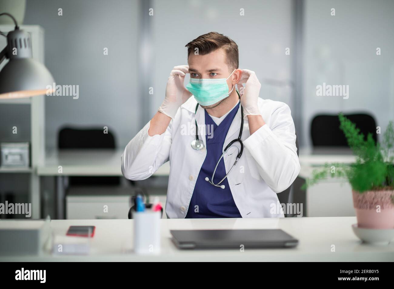 A young therapist in a medical mask sits in his office in a private ...