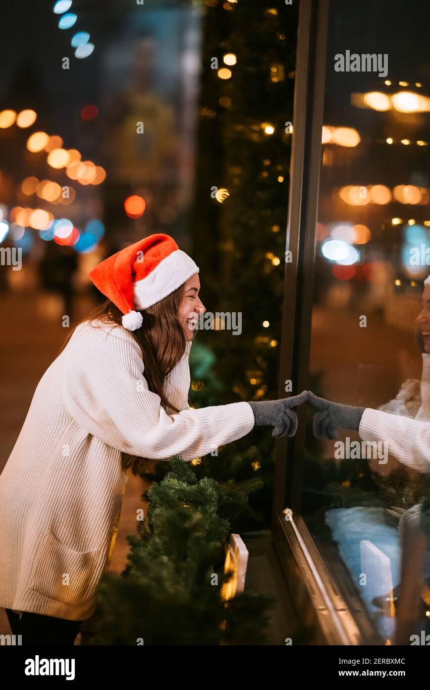 Smiling girl looking through shop window, city lights on background ...