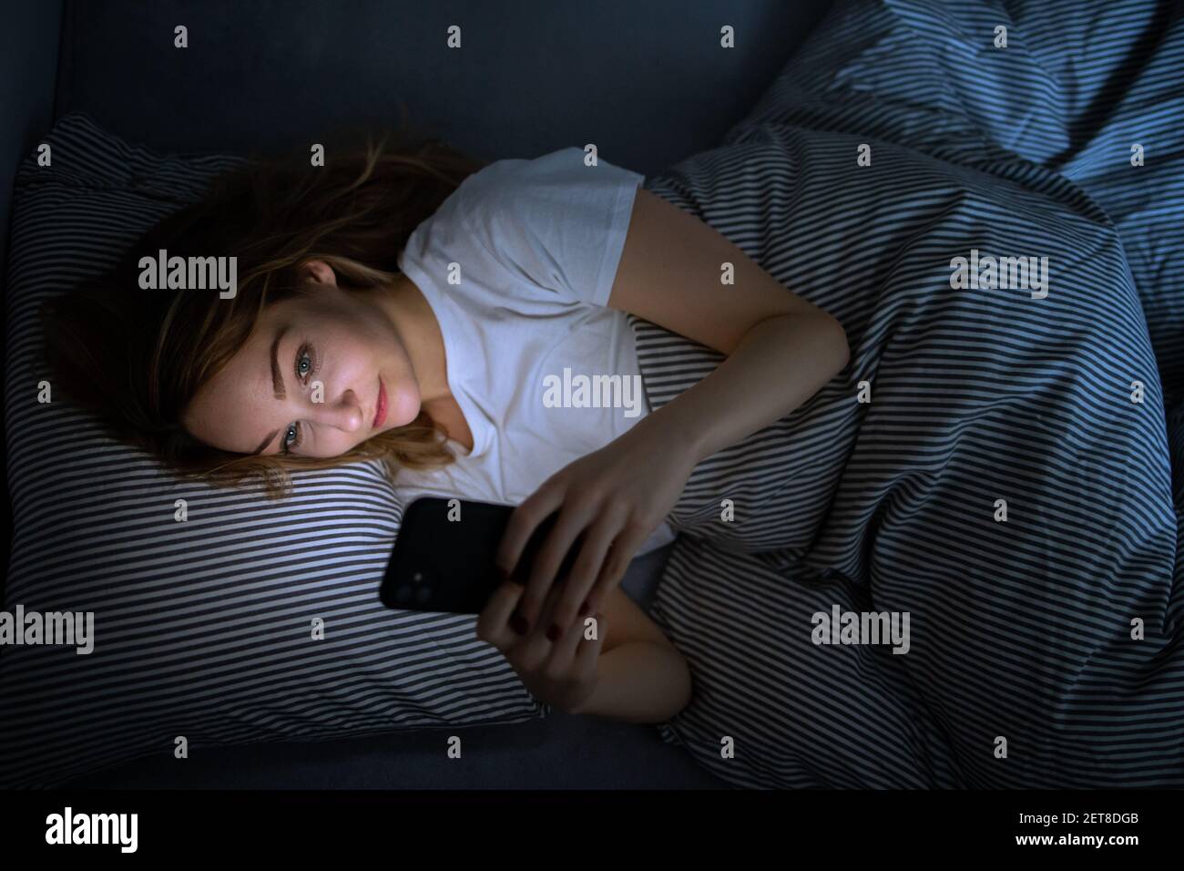 Young woman in bed holding a phone, tired and exhausted, blue light ...