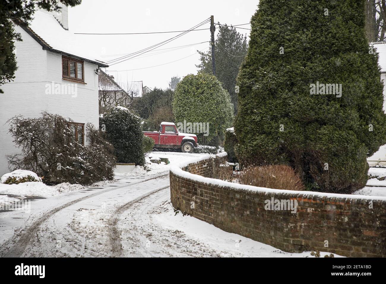 A car trails on a snow pathway with a white building and a red truck ...