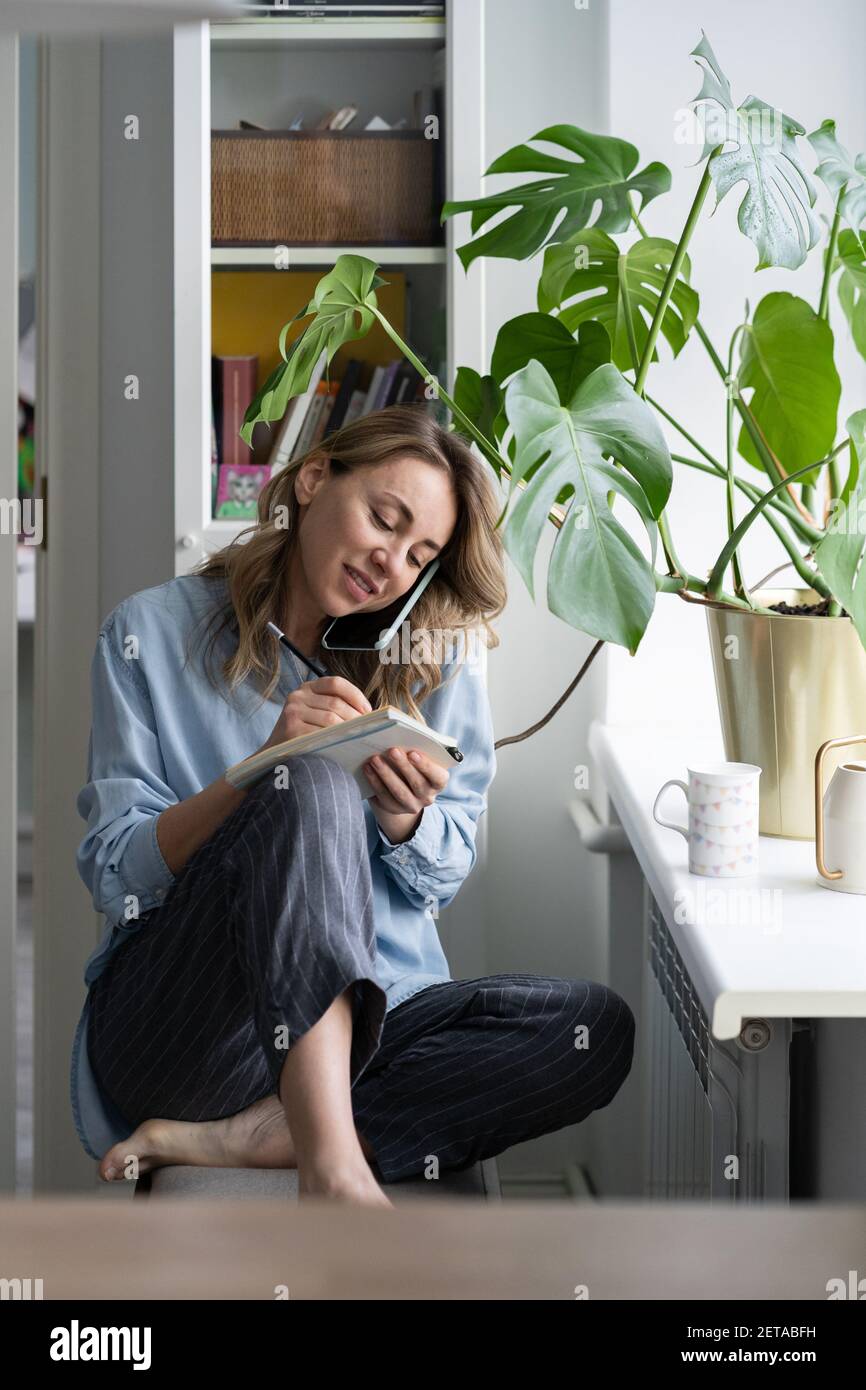Smiling woman talking on cellphone writes in a notebook, sitting next ...