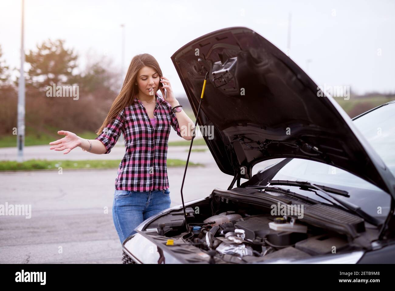 Young worried girl is using a phone to call the mechanic to fix the car whos hood she is looking ...