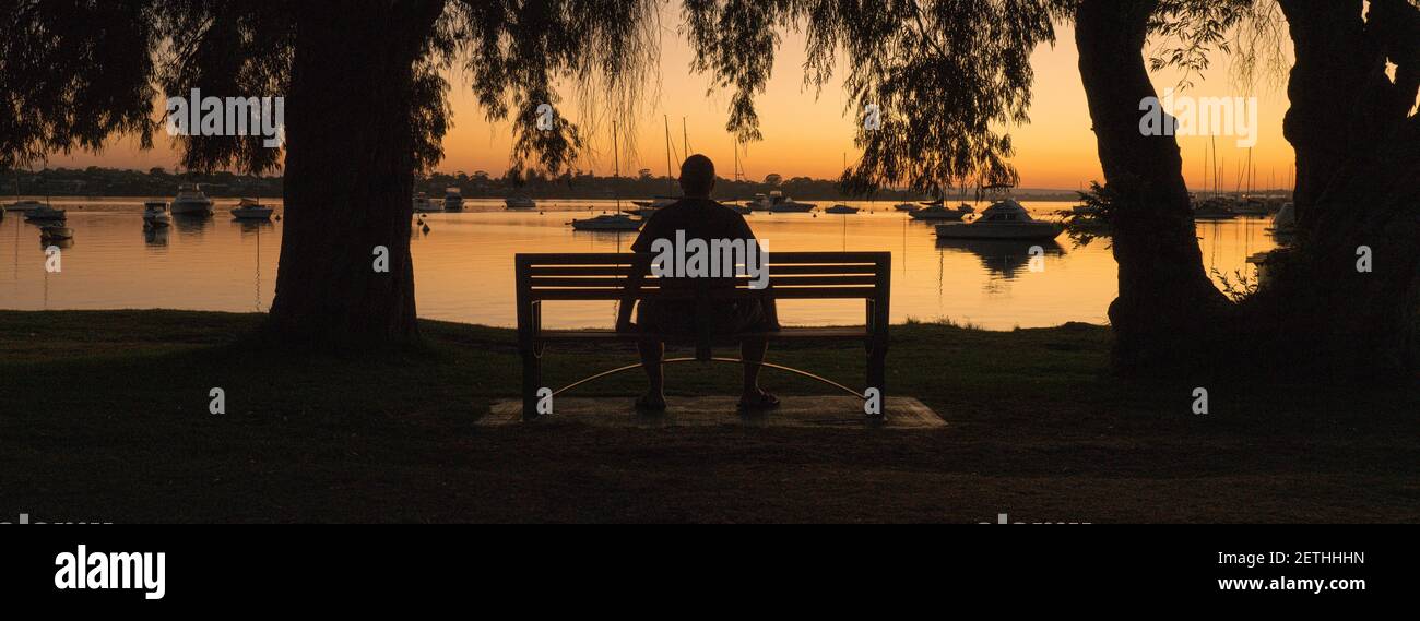 A man sitting on a beach near a shore watching a beautiful sunset in ...