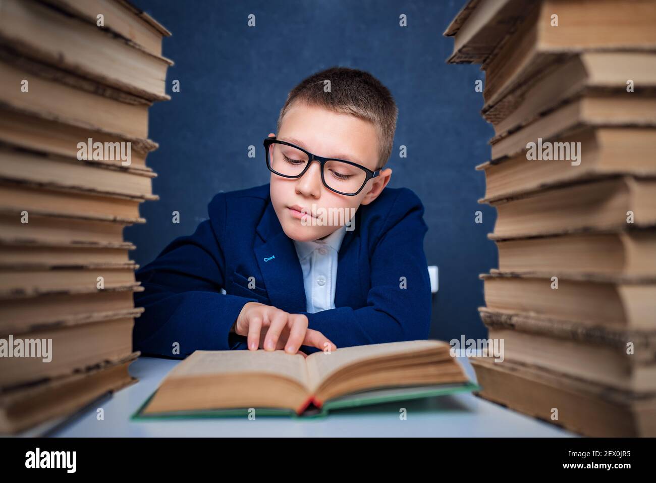 Smart boy in glasses sitting between two piles of books and read book ...