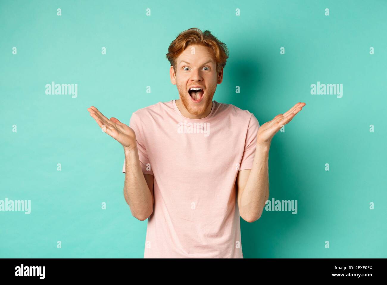 Portrait of surprised and happy young man with red hair, spread hands ...