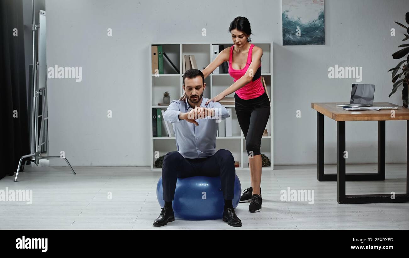 Fitness trainer standing near businessman on fitness ball in office ...
