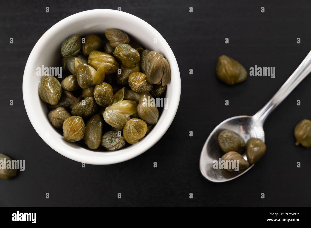 Top view of a bowl of green capers and a spoon isolated on a black ...