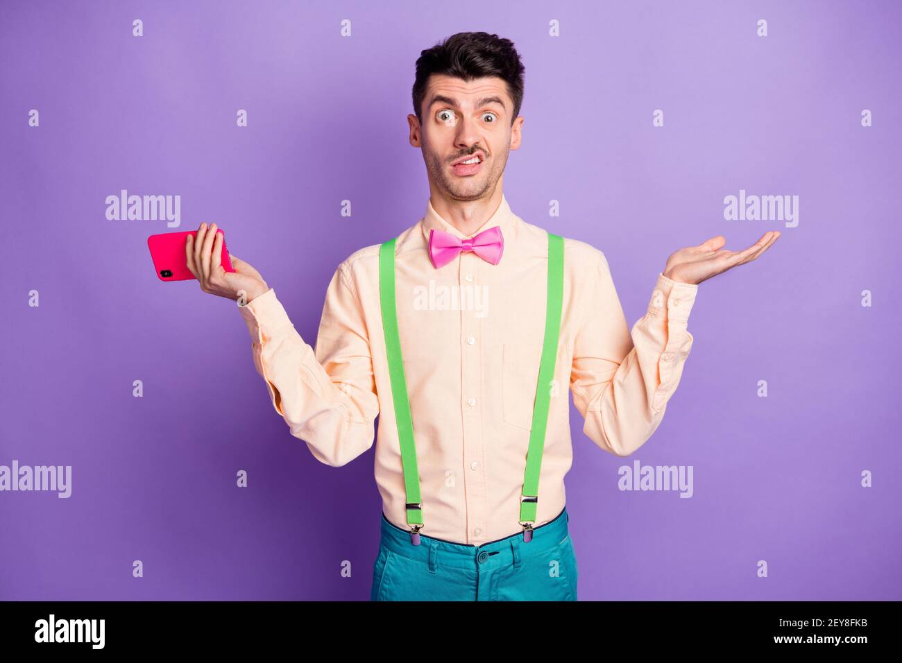 Photo of uncertain young man raise hands clueless hold smartphone wear ...