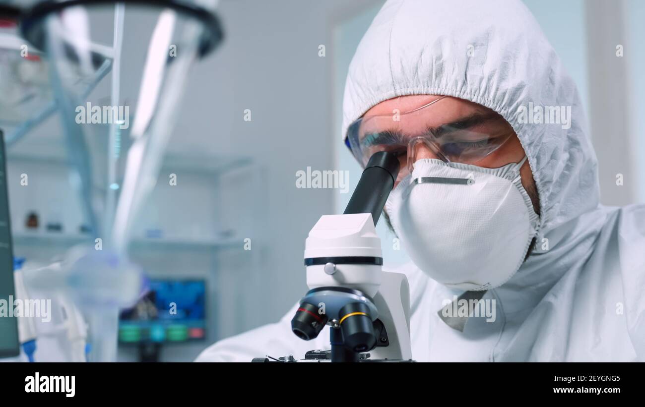 Biochemist in ppe suit looking through a microscope during experiment ...