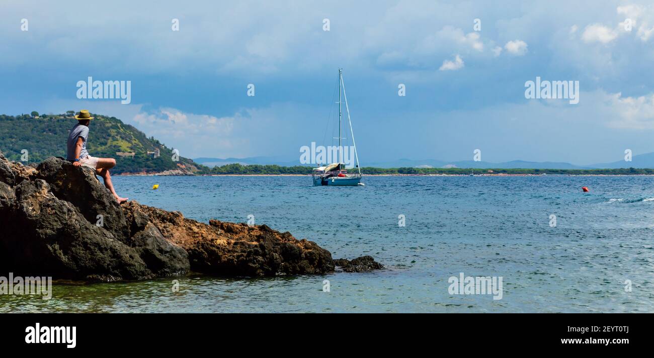 A man sitting on rocks looks over the horizon and the coast. A boat is ...