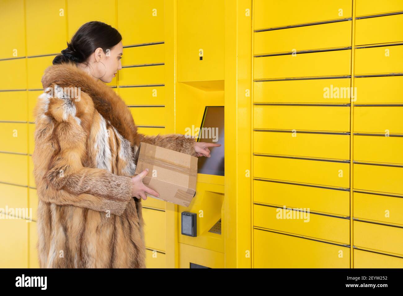 A young adult in a fur coat operates the touch panel of a parcel locker ...