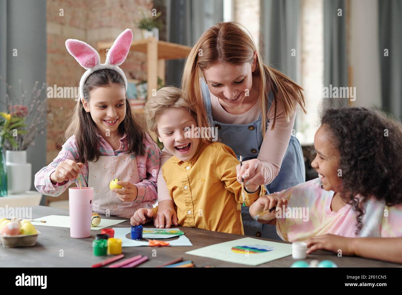 Young cheerful woman bending over table with group of happy kids while ...