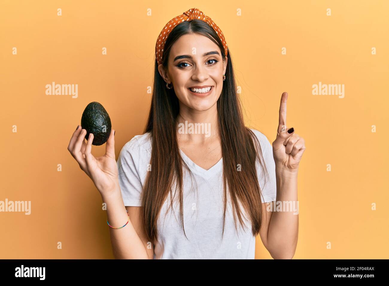 Beautiful brunette young woman holding avocado smiling with an idea or ...