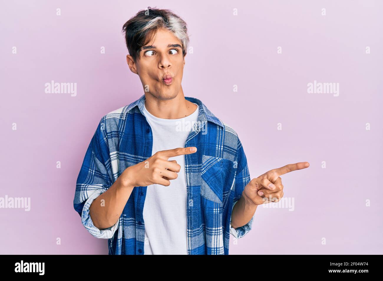 Young hispanic man pointing with fingers to the side making fish face ...