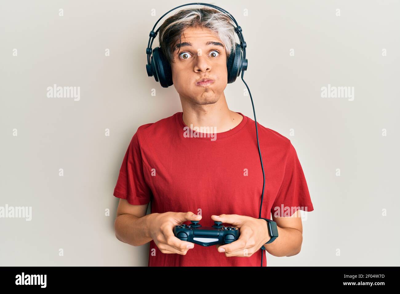 Young hispanic man playing video game holding controller puffing cheeks ...