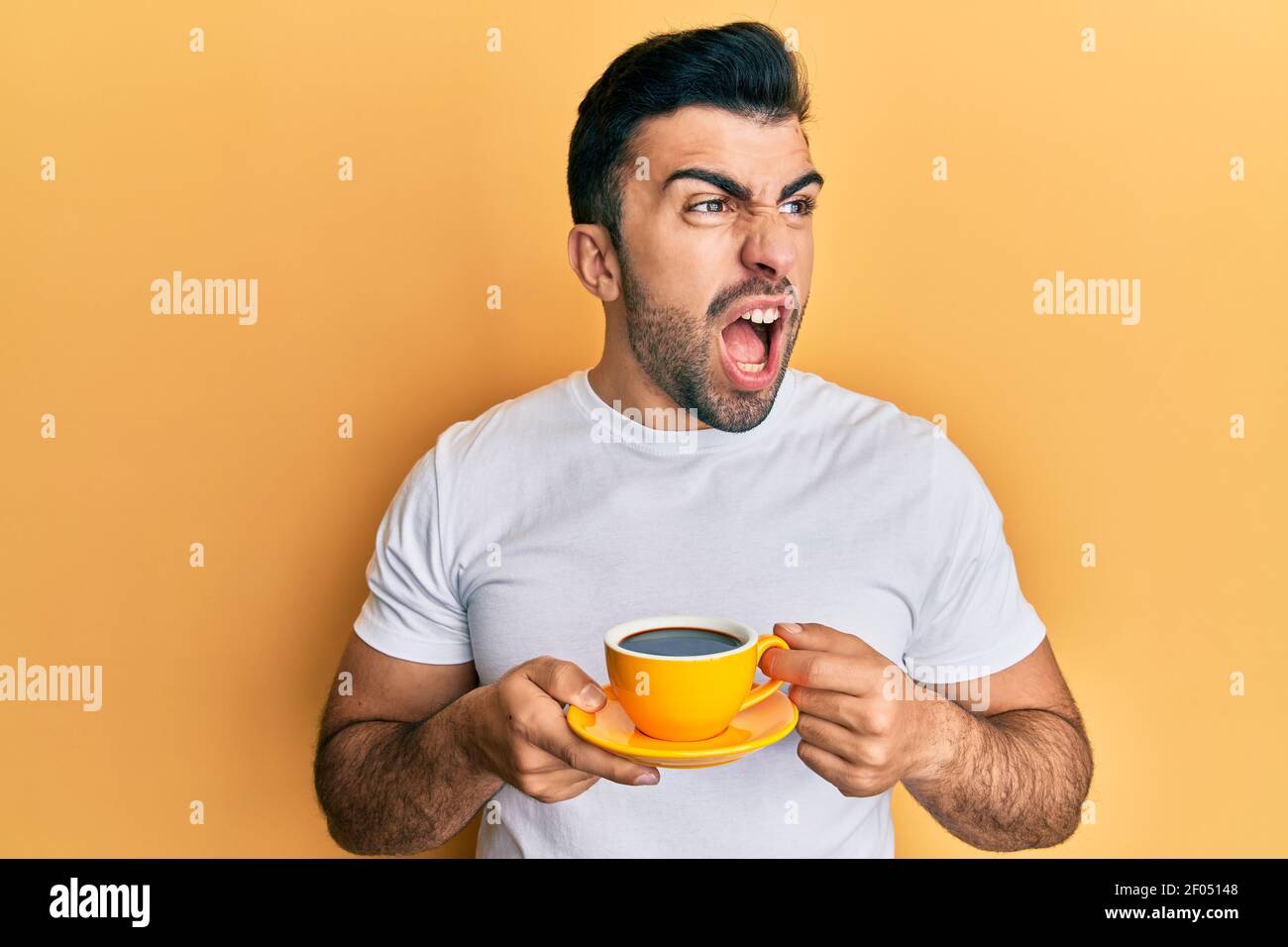 Young hispanic man drinking a cup of coffee angry and mad screaming ...