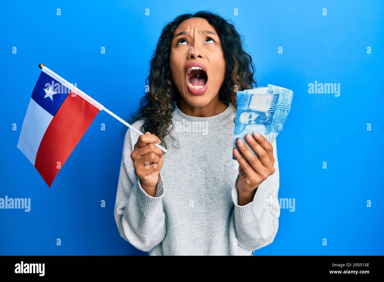 Young latin woman holding chile flag and chilean pesos banknotes angry ...
