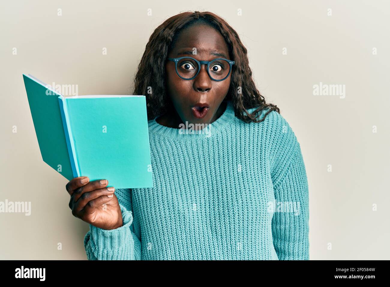 African young woman reading a book wearing glasses scared and amazed ...