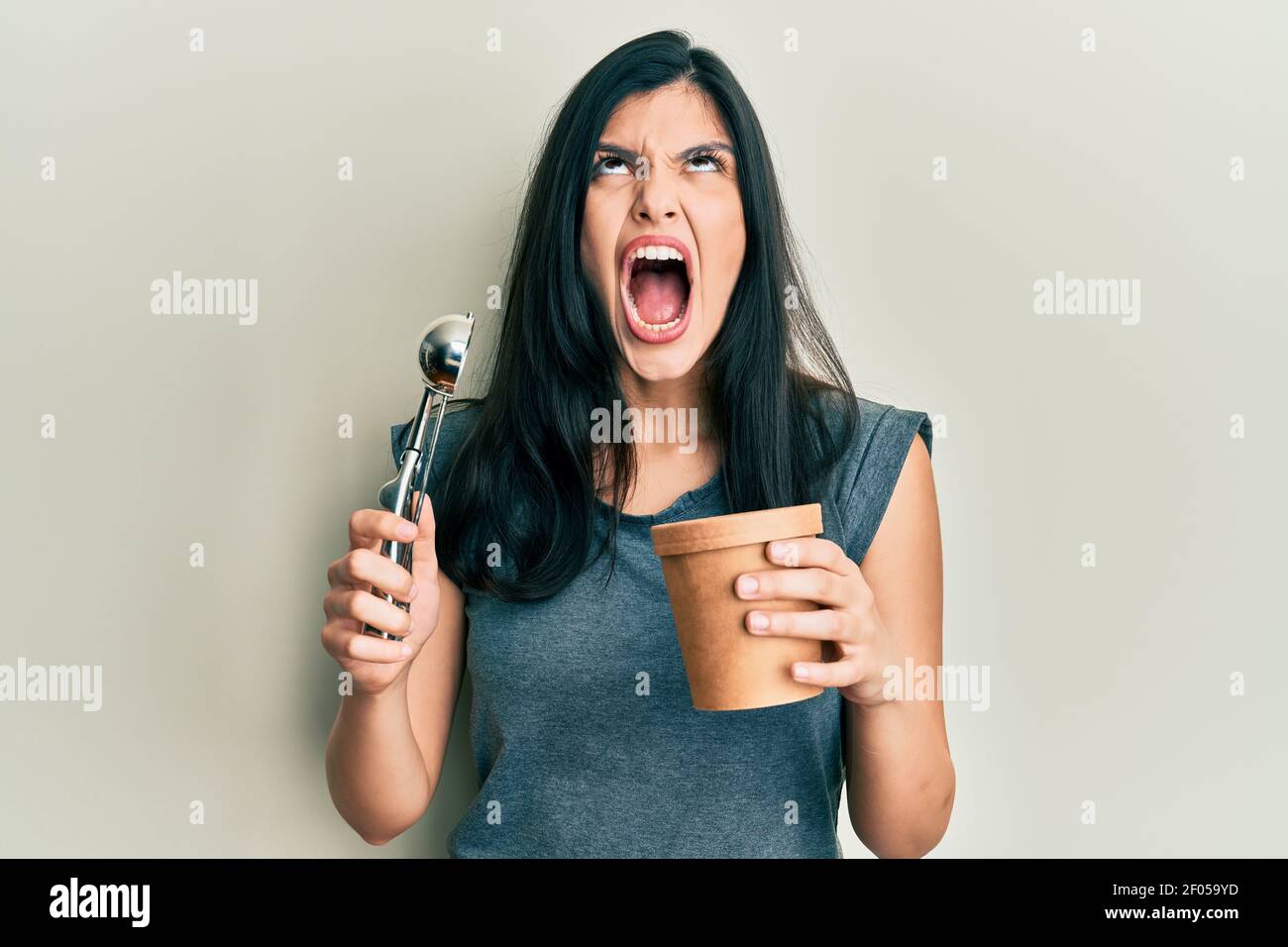 Young hispanic woman holding ice cream angry and mad screaming ...