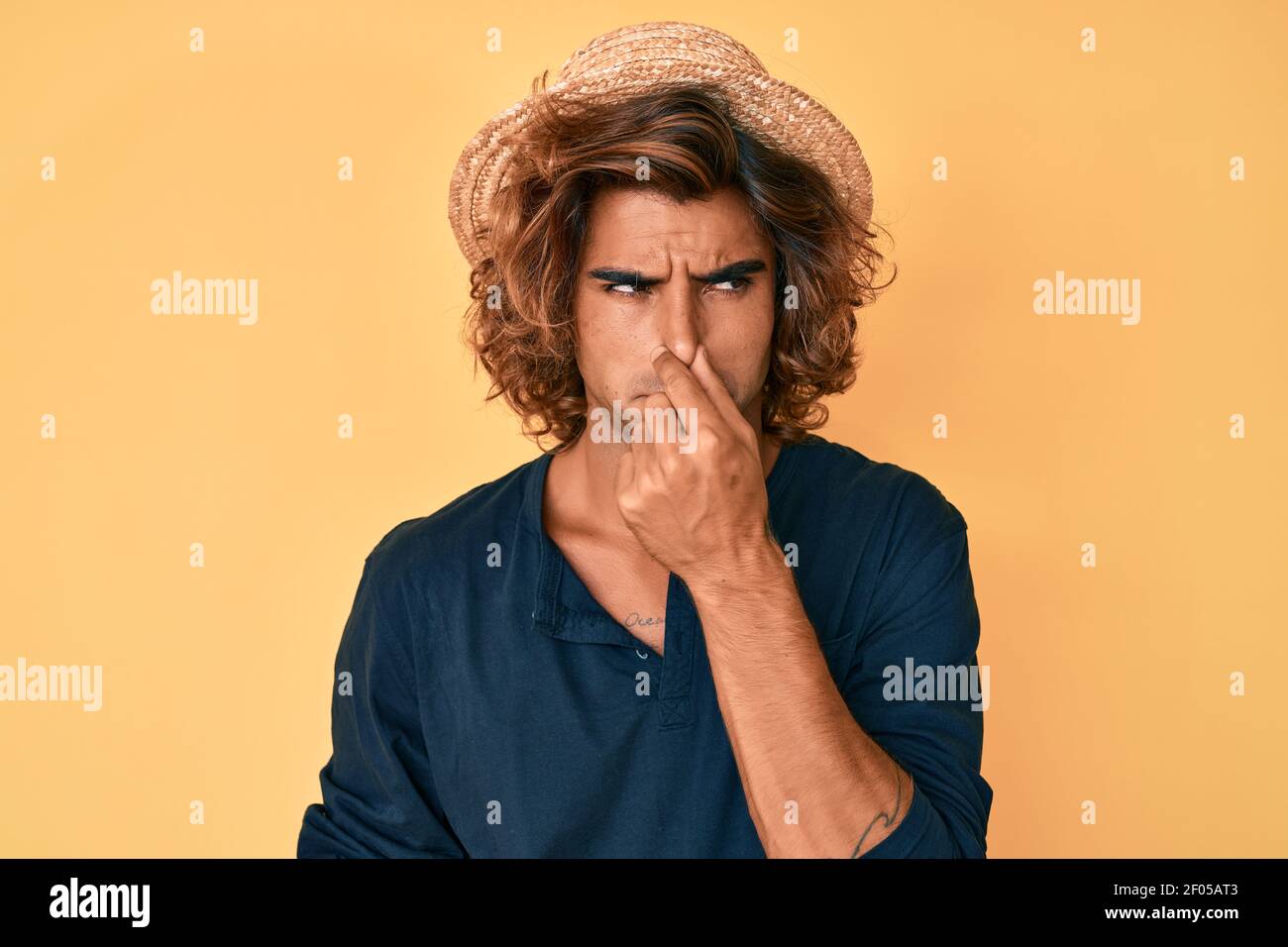 Young hispanic man wearing summer hat smelling something stinky and ...