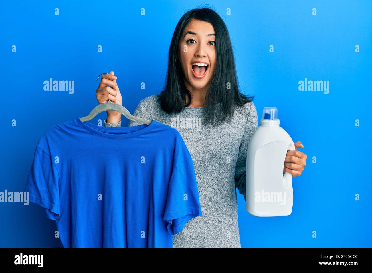 Beautiful asian young woman holding blue t shirt and detergent bottle celebrating crazy and ...