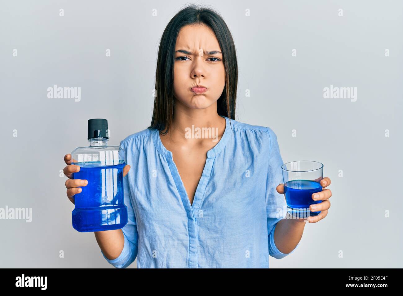 Young hispanic girl holding mouthwash for fresh breath puffing cheeks ...
