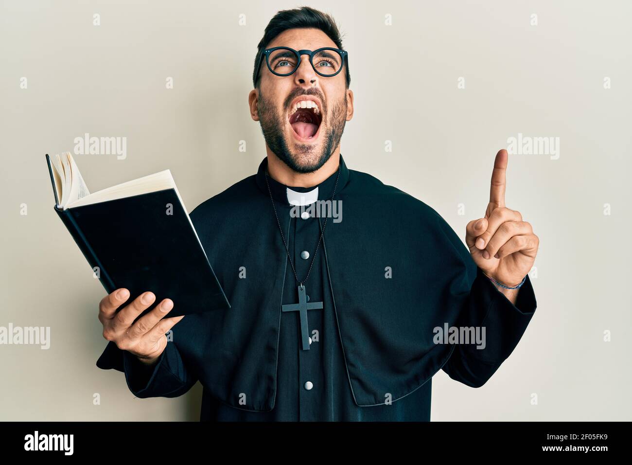 Young hispanic priest man holding bible with finger up angry and mad ...